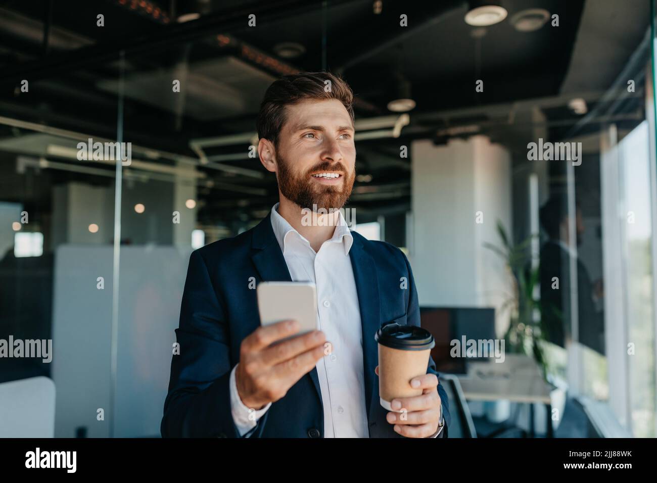 Happy handsome businessman in suit with cup of coffee using smartphone ...