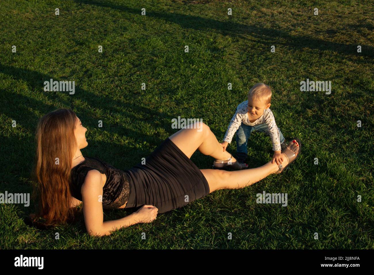 Little toddler son playing with mother legs, woman with her child outdoor on the grass lawn ...