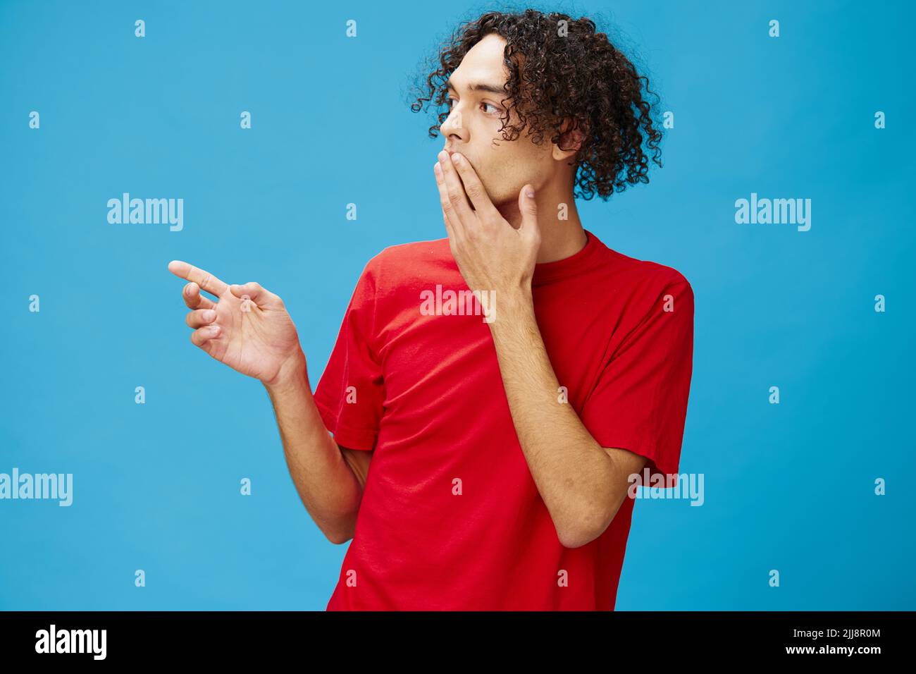Excited shocked curly tanned Caucasian guy in basic red t-shirt point finger at free place looks ...