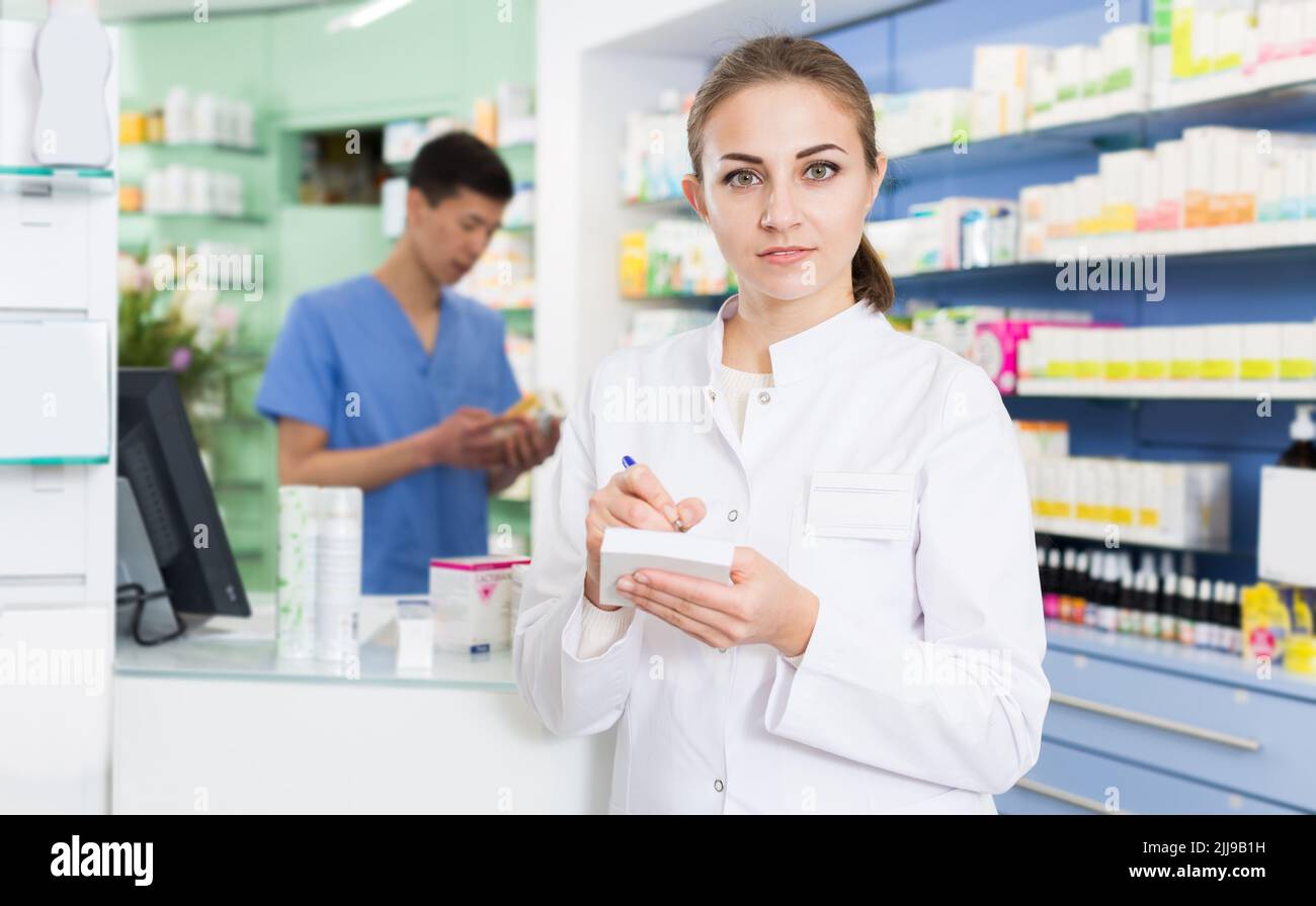 Young female pharmacist is attentively checking medicine to notebook ...
