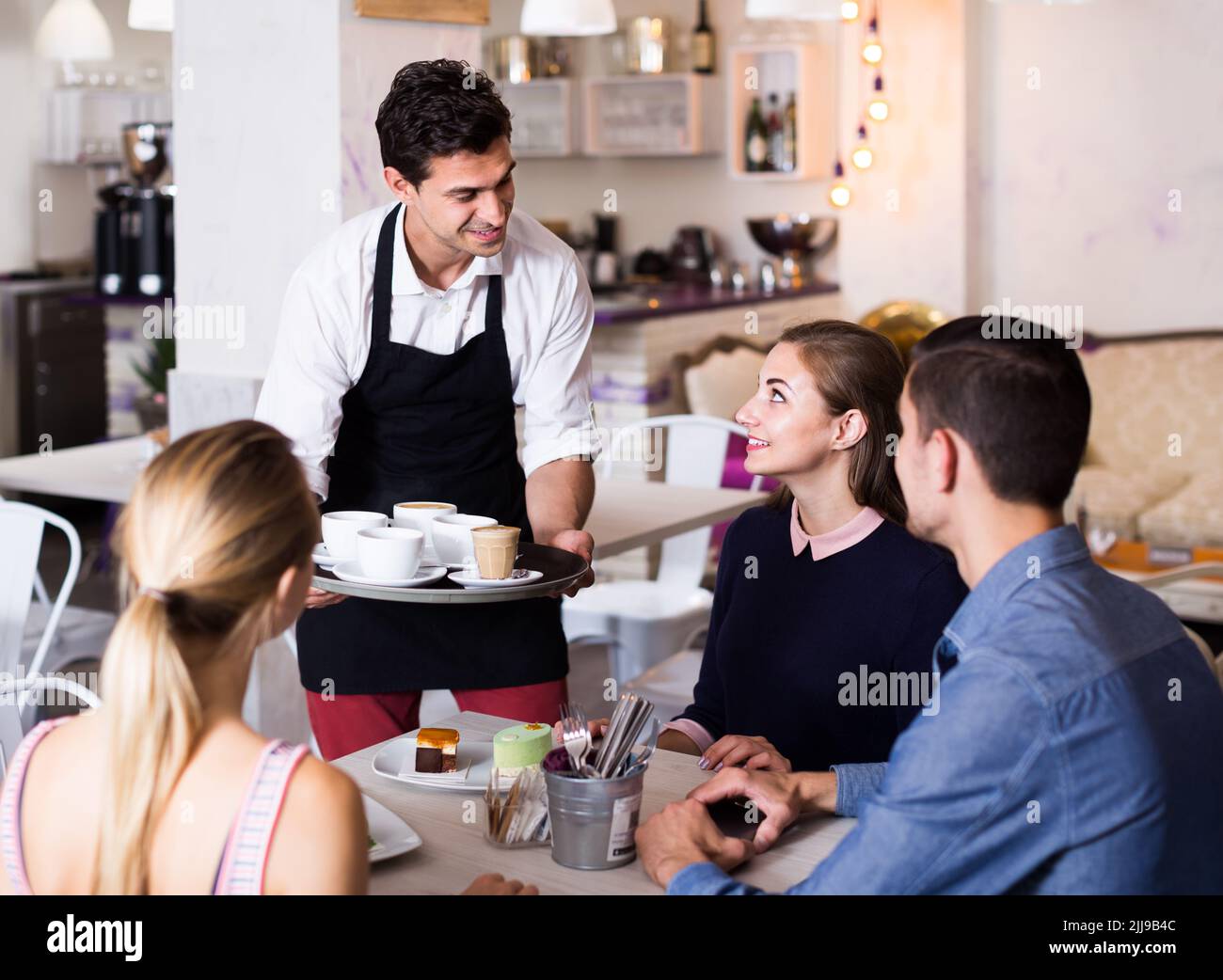Polite smiling waiter bringing ordered dishes to friends in tearoom ...
