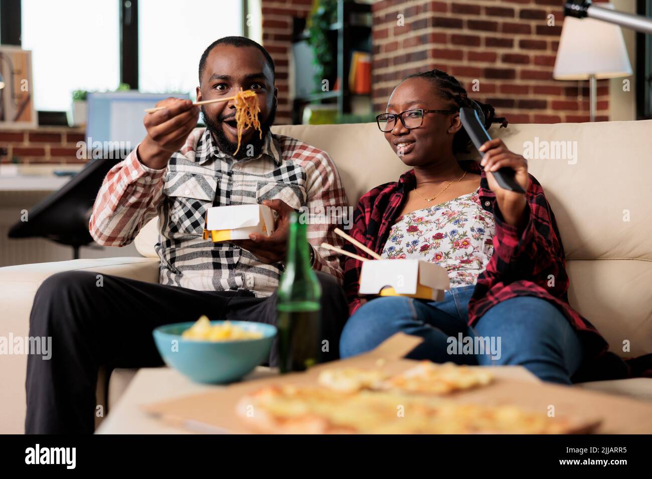 Man eating takeaway noodles with chopsticks and woman using remote ...