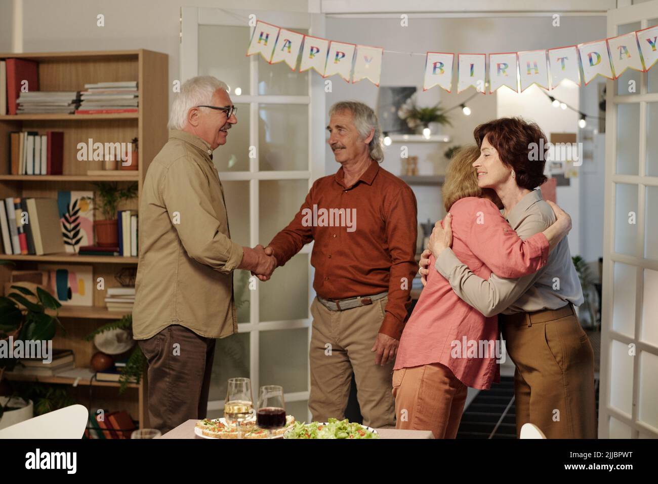 Two senior women giving each other hug while their husbands shaking ...