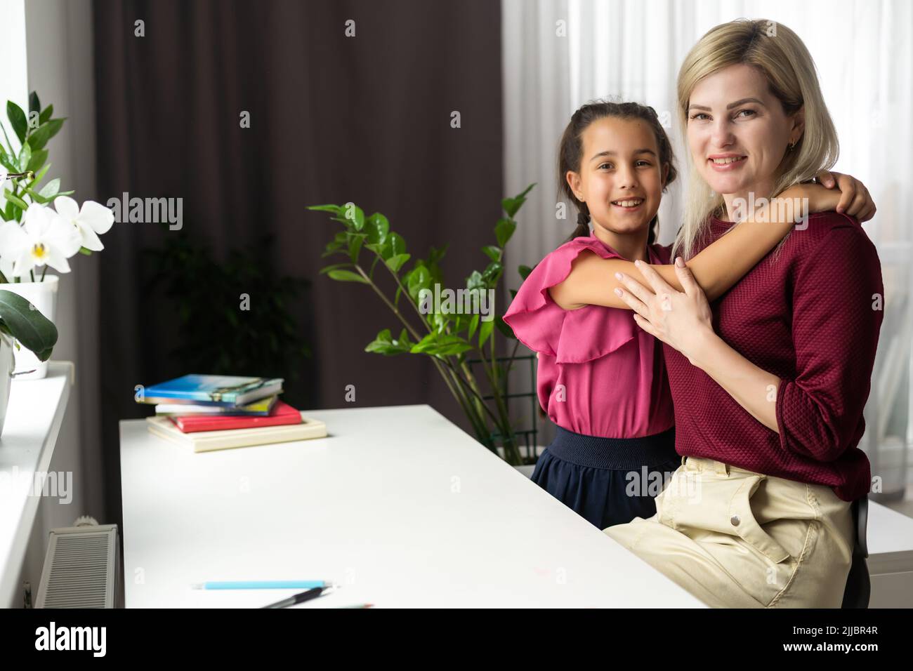 Caring mother calming and hugging little daughter, sitting at desk ...