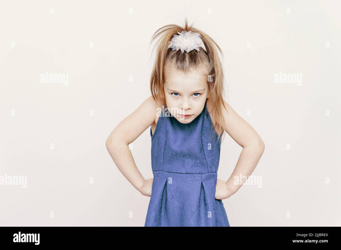portrait of grumpy little cute girl in blue dress. hands to the sides ...