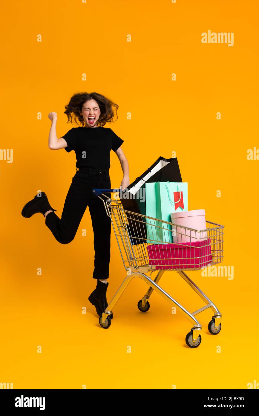 Excited woman making winner gesture while posing with shopping cart ...