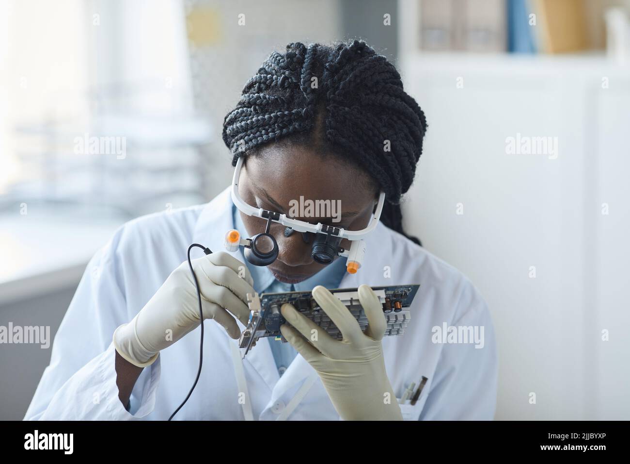 Portrait of young black woman inspecting computer parts and wearing magnifying visor while ...