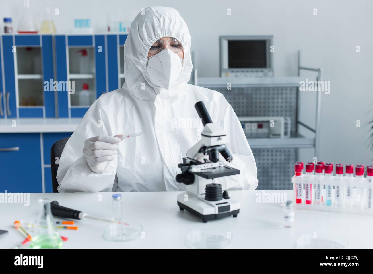 Scientist in hazmat suit holding glass near microscope and test tubes ...