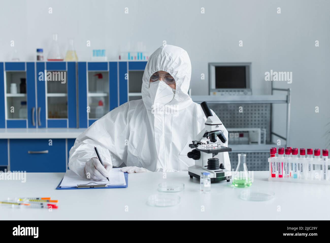 Scientist in hazmat suit writing on clipboard near microscope and test ...