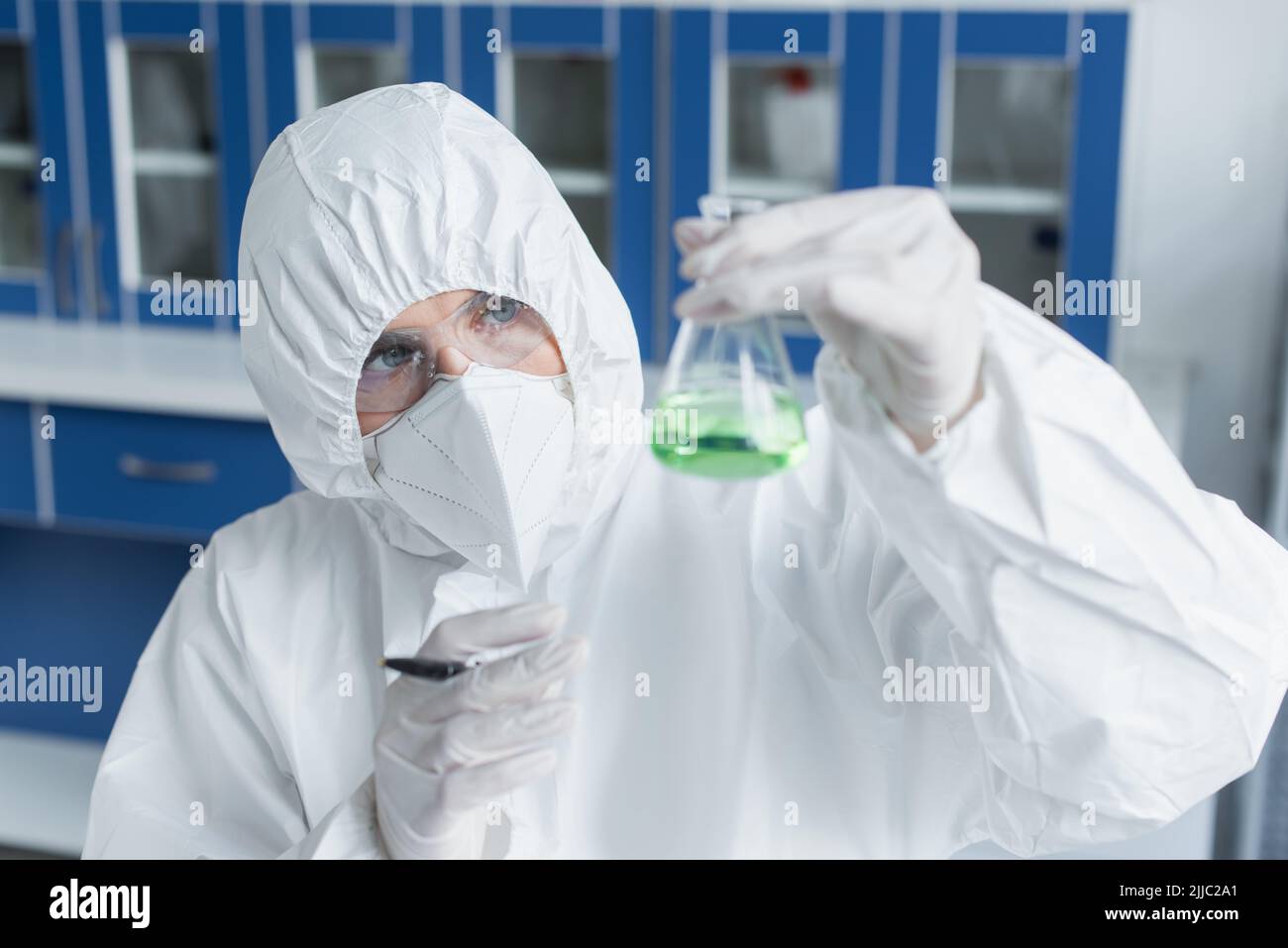 Scientist in protective suit and mask holding flask with liquid in lab ...