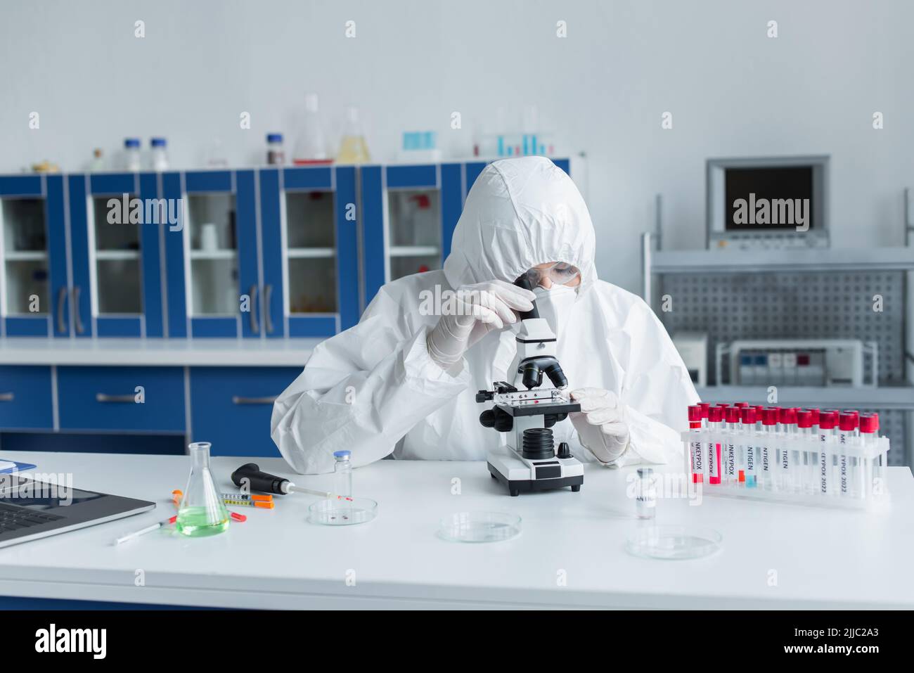 Scientist in hazmat suit using microscope near test tubes with ...