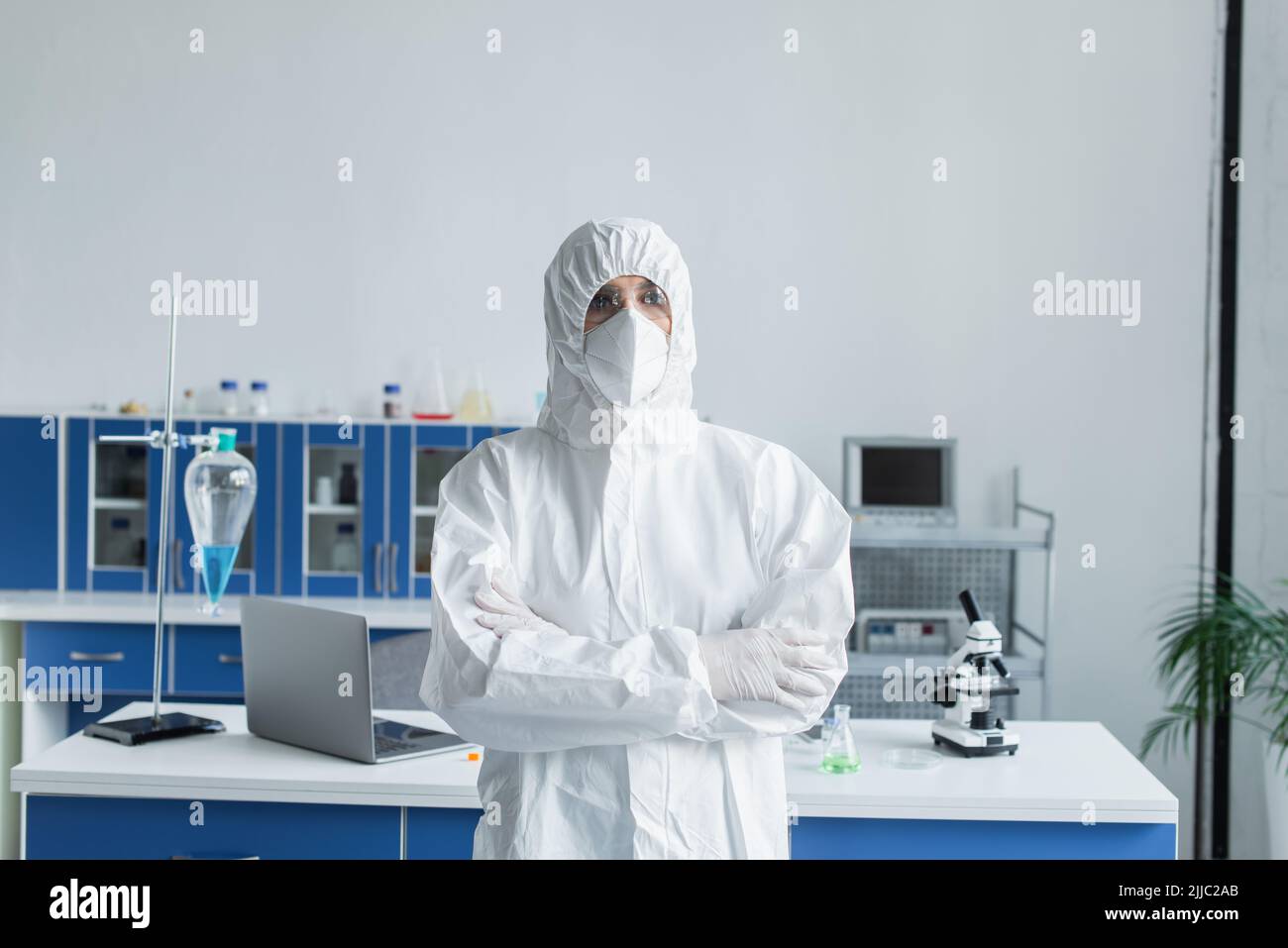 Scientist in hazmat suit and protective goggles crossing arms in lab ...