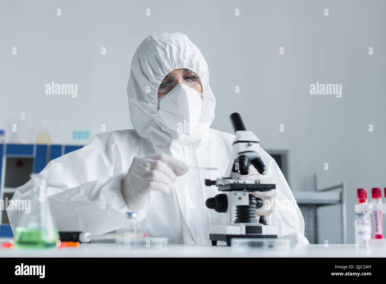 Scientist in hazmat suit holding glass while working with microscope in ...
