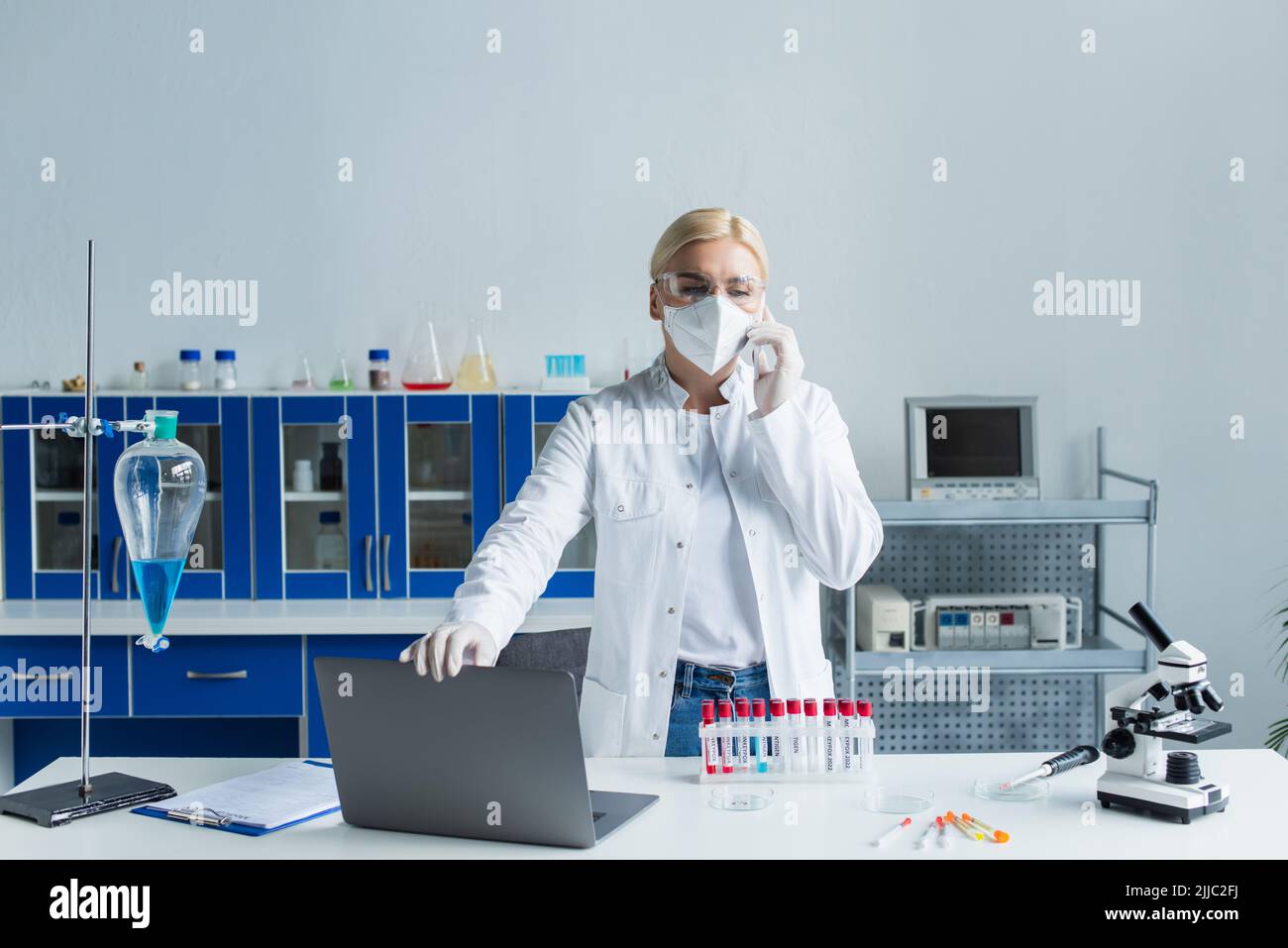 Scientist in goggles and mask talking on smartphone near test tubes and ...