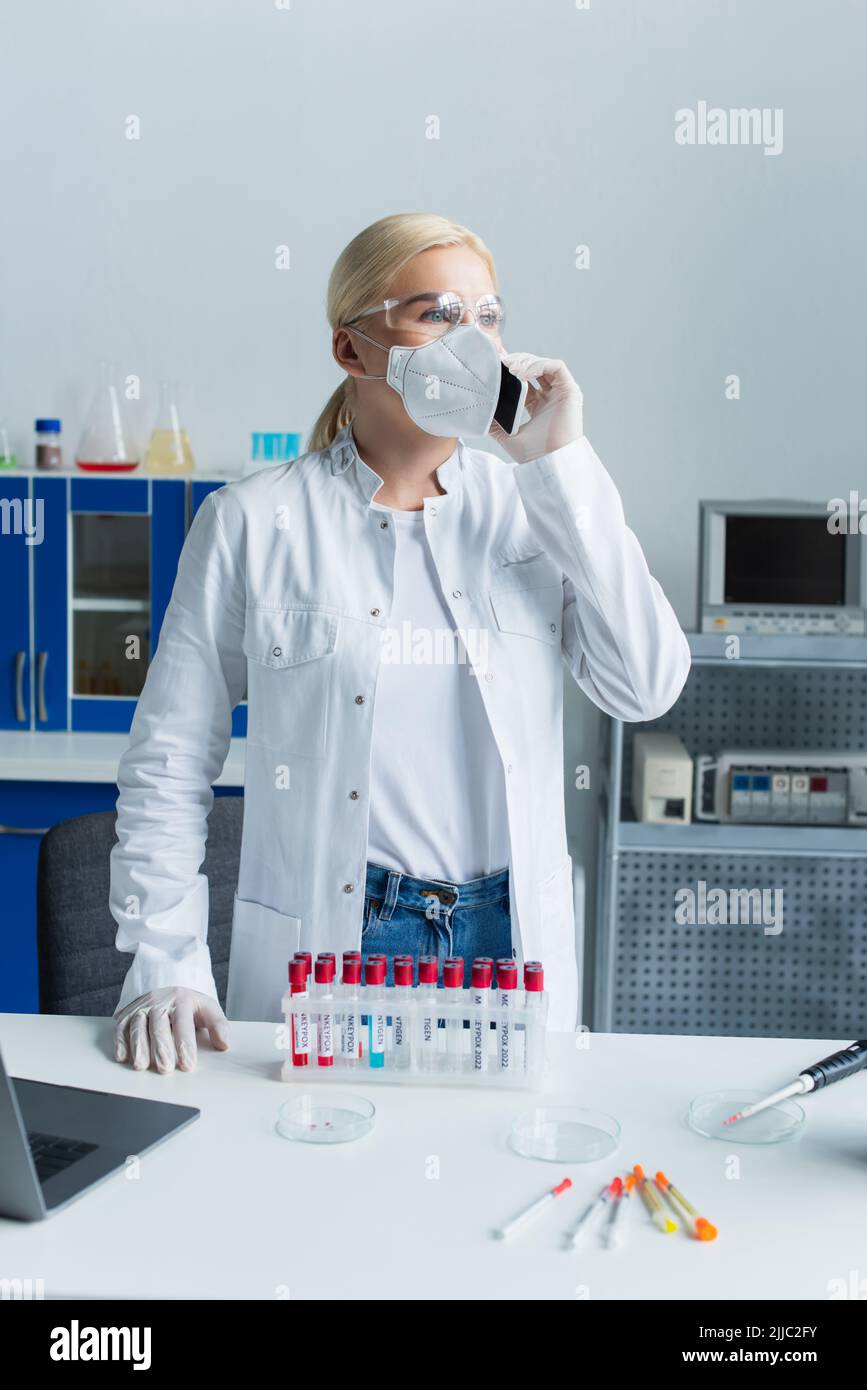 Researcher in medical mask talking on smartphone near test tubes and ...