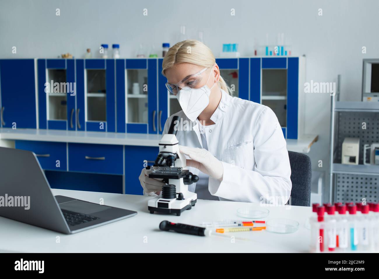 Scientist in protective mask using microscope near test tubes in ...