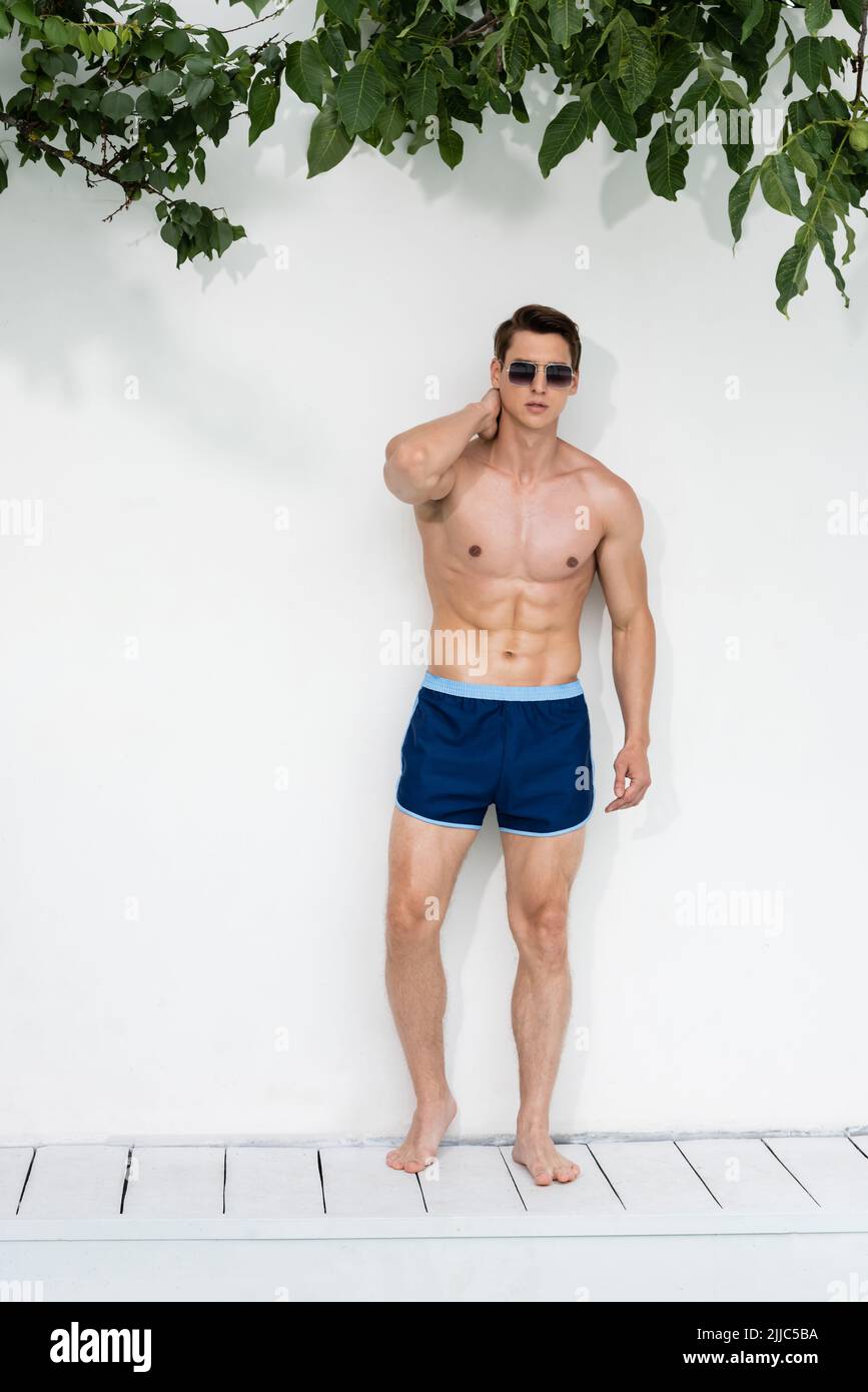 full length of athletic man in swimming trunks and sunglasses posing ...