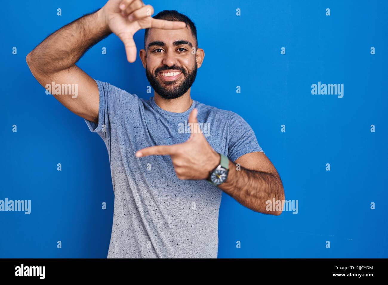 Middle east man with beard standing over blue background smiling making ...