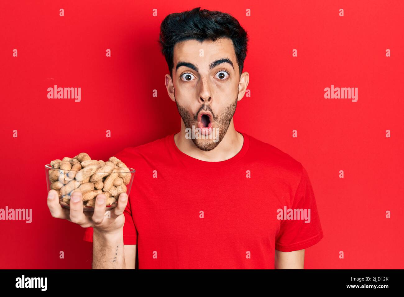 Young hispanic man holding peanuts scared and amazed with open mouth ...
