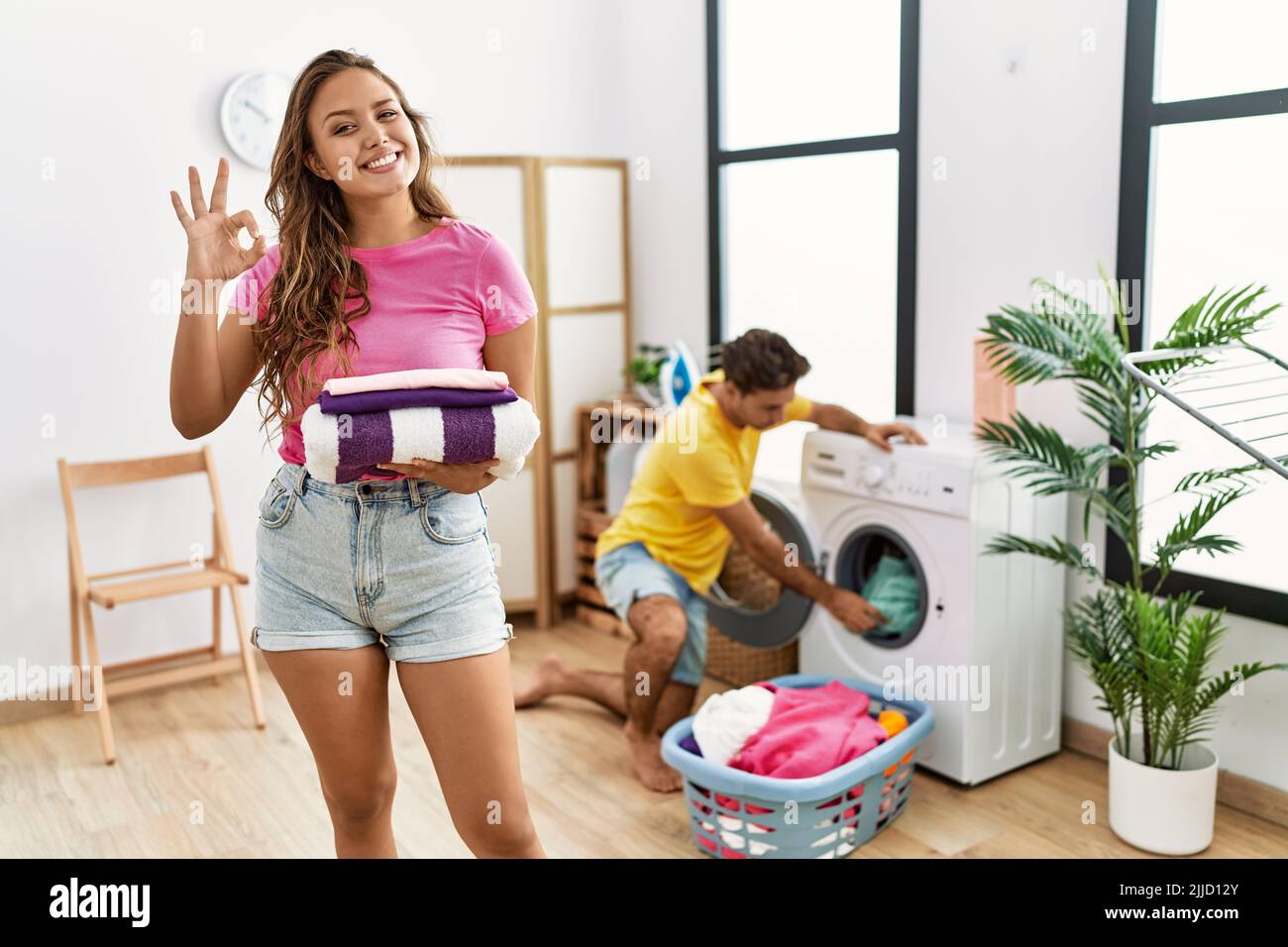 Young hispanic couple putting dirty laundry into washing machine doing ok sign with fingers ...