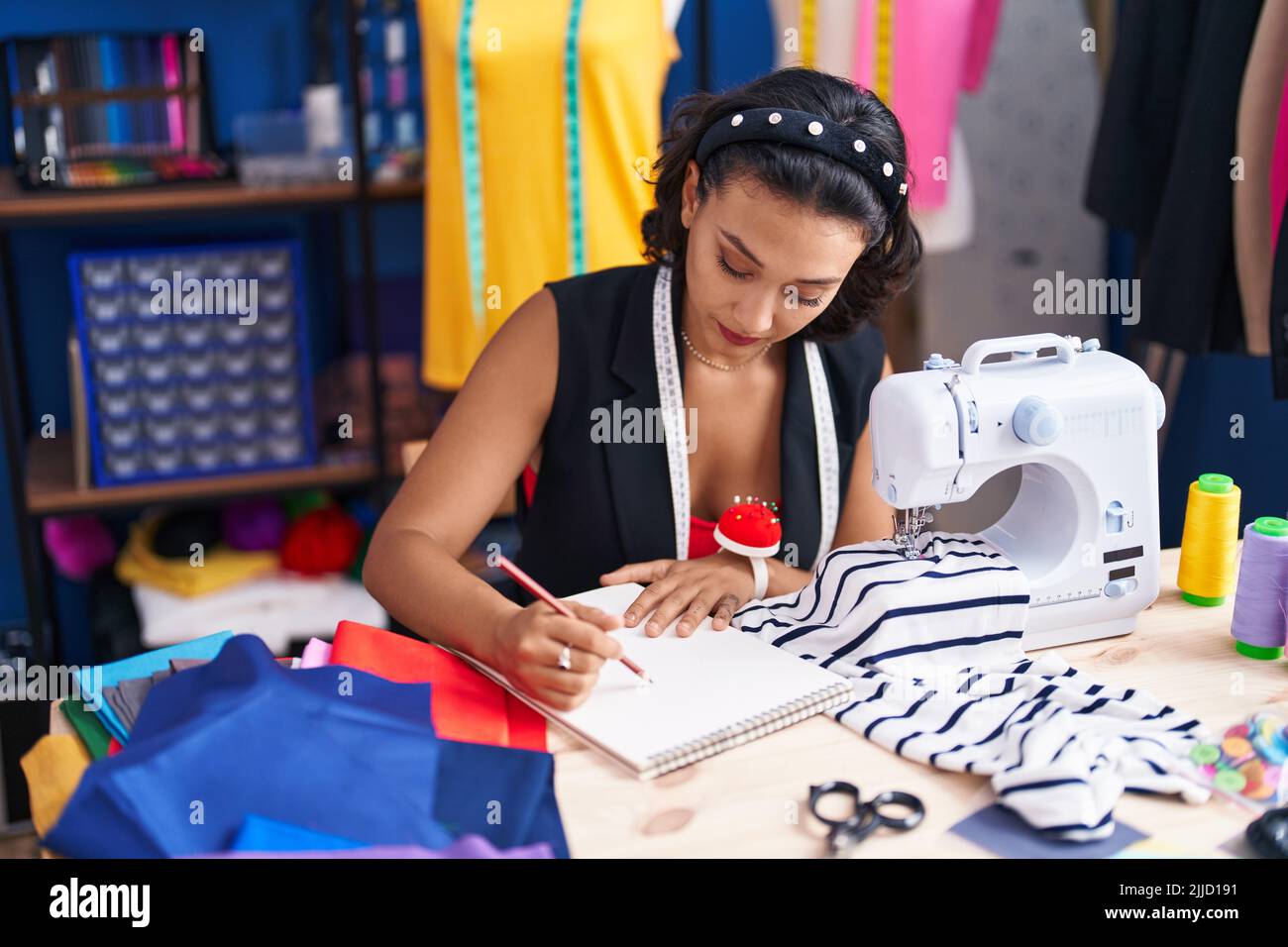 Young beautiful hispanic woman tailor drawing on notebook at clothing ...