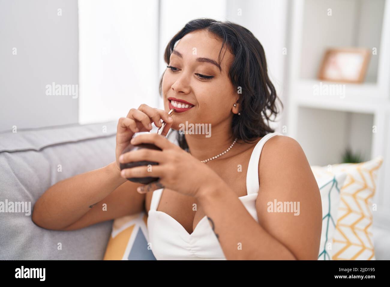 Young beautiful hispanic woman drinking mate infusion sitting on sofa ...