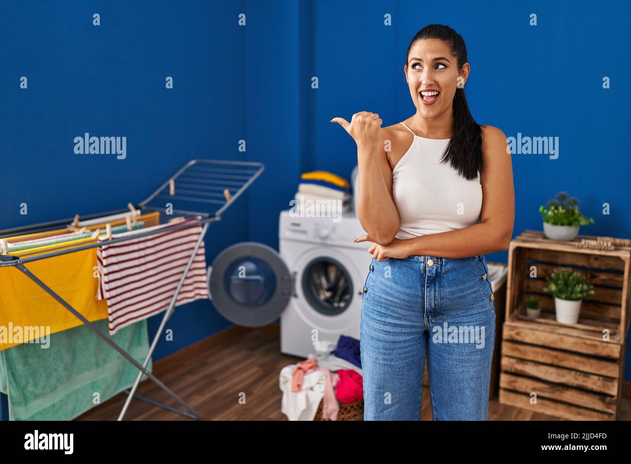 Young hispanic woman at laundry room smiling with happy face looking and pointing to the side ...