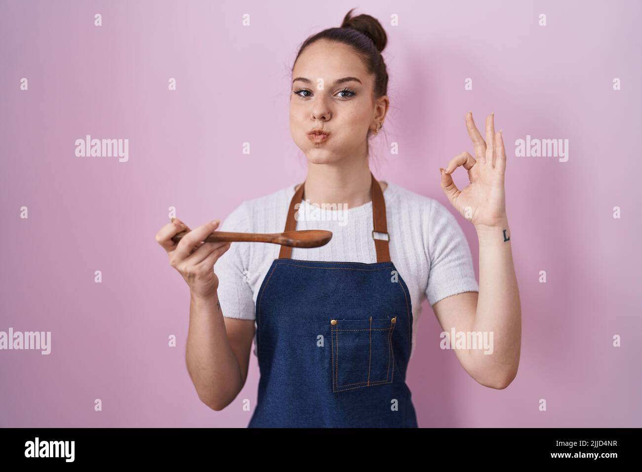 Young hispanic girl wearing professional cook apron holding wood spoon ...