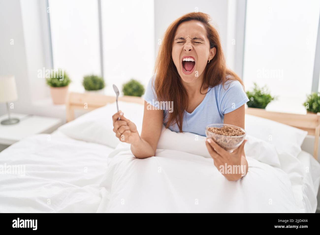 Brunette woman eating breakfast in the bed angry and mad screaming ...