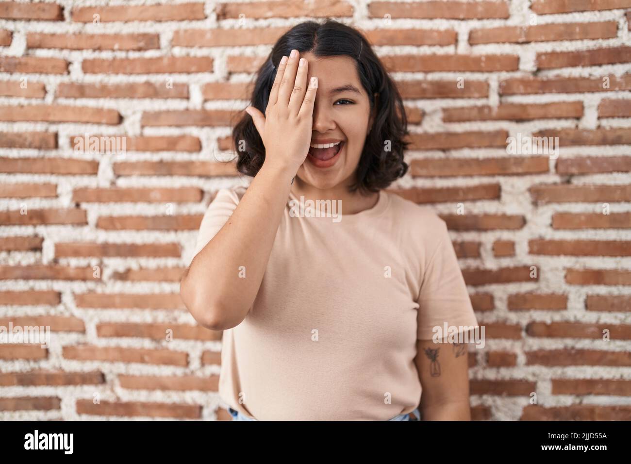 Young hispanic woman standing over bricks wall covering one eye with ...