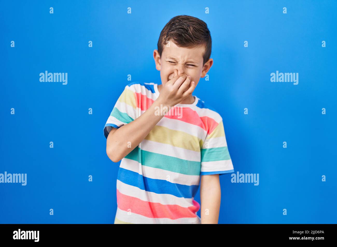 Young caucasian kid standing over blue background smelling something ...