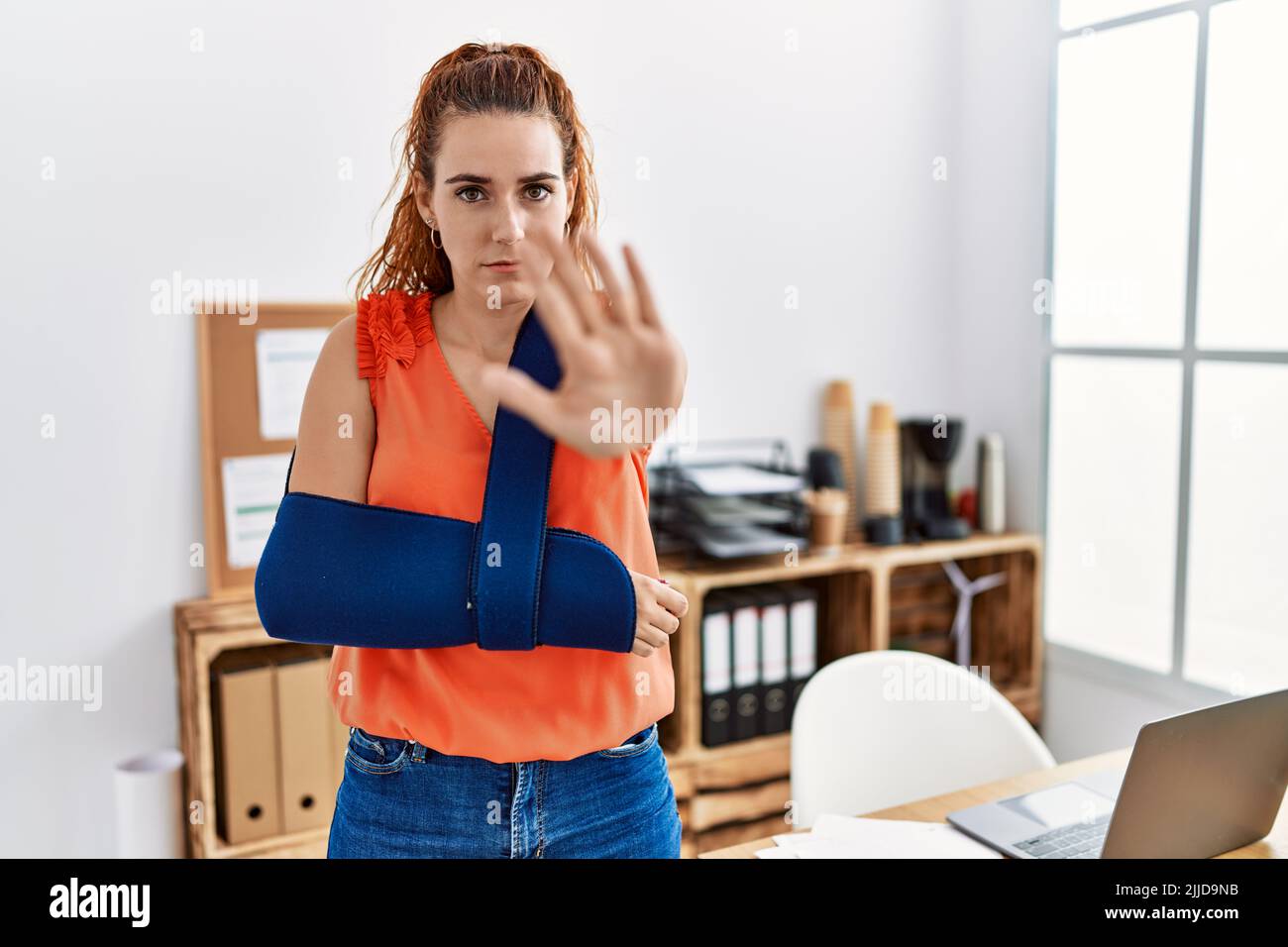 Young redhead woman wearing arm on sling at the office with open hand ...