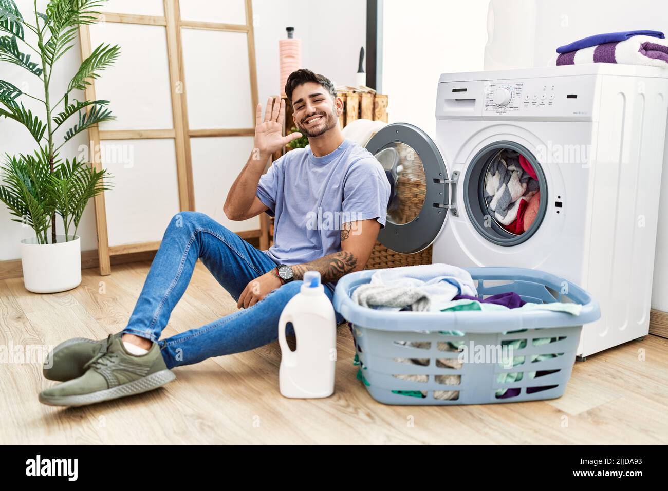 Young hispanic man putting dirty laundry into washing machine waiving ...