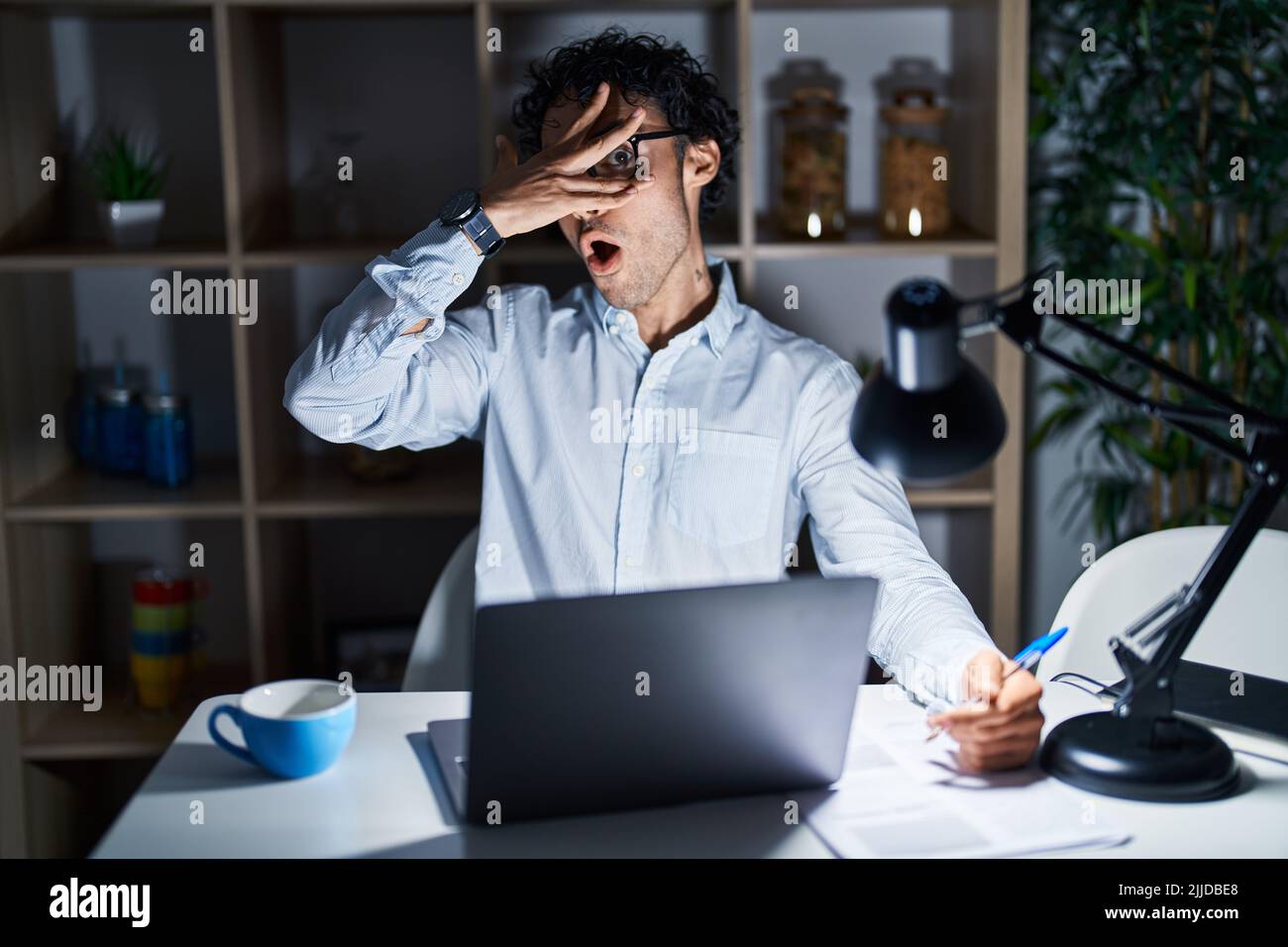 Hispanic man working at the office at night peeking in shock covering ...