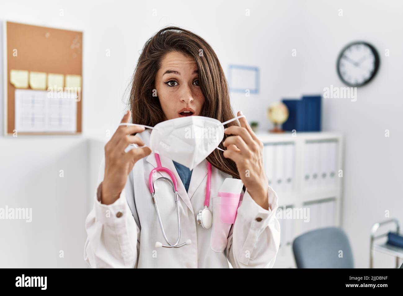 Young doctor woman wearing safety mask at the clinic in shock face ...