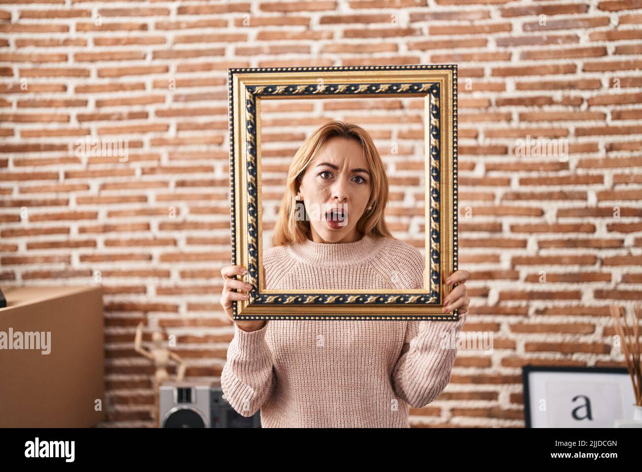 Hispanic woman holding empty frame in shock face, looking skeptical and ...