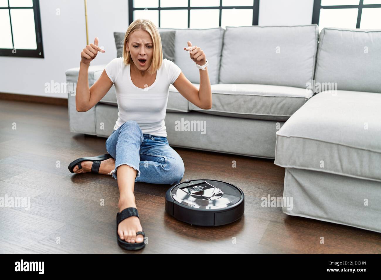 Young caucasian woman sitting at home by vacuum robot pointing down ...