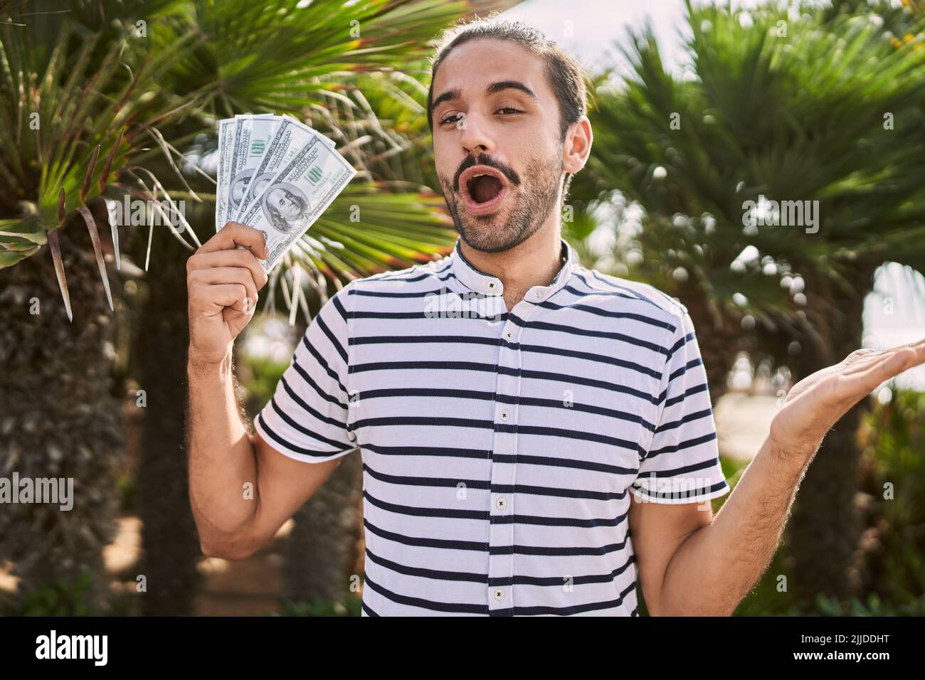 Young hispanic man holding dollars close to face celebrating ...