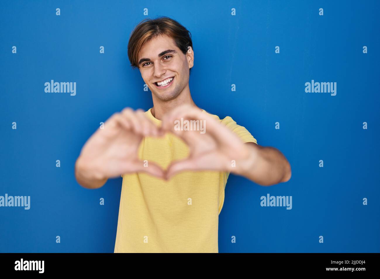Young man standing over blue background smiling in love doing heart ...