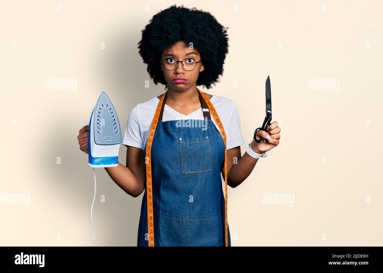 Young african american woman dressmaker designer wearing atelier apron holding iron and scissors ...