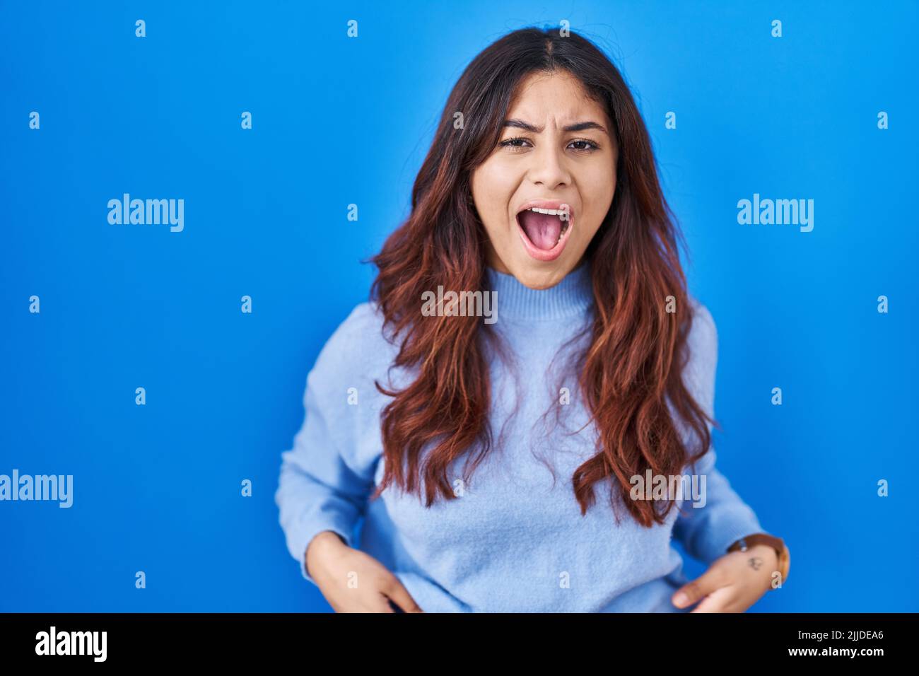 Hispanic young woman standing over blue background angry and mad ...