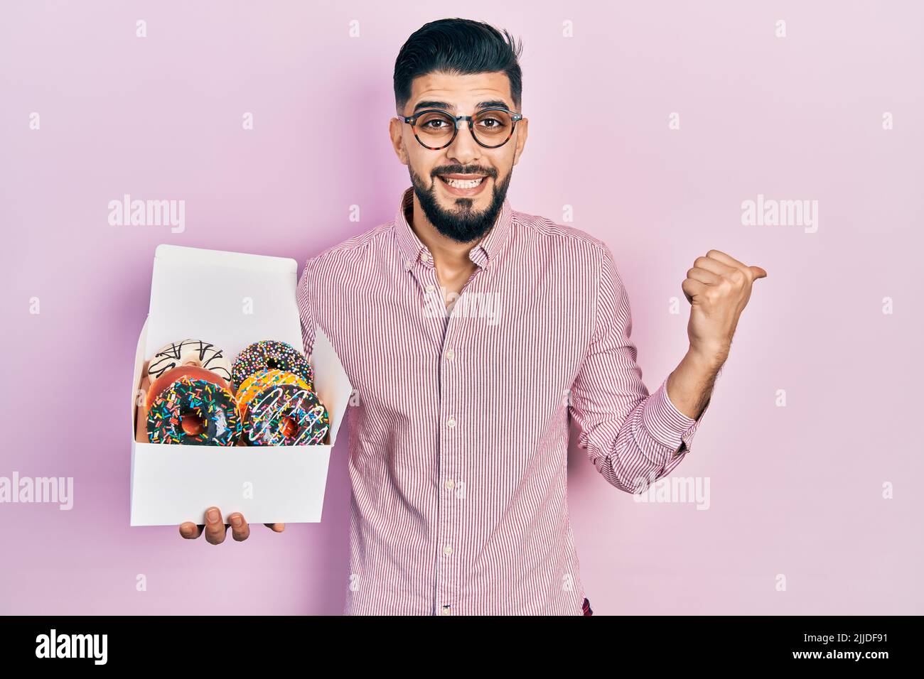 Handsome man with beard holding tasty colorful doughnuts pointing thumb ...