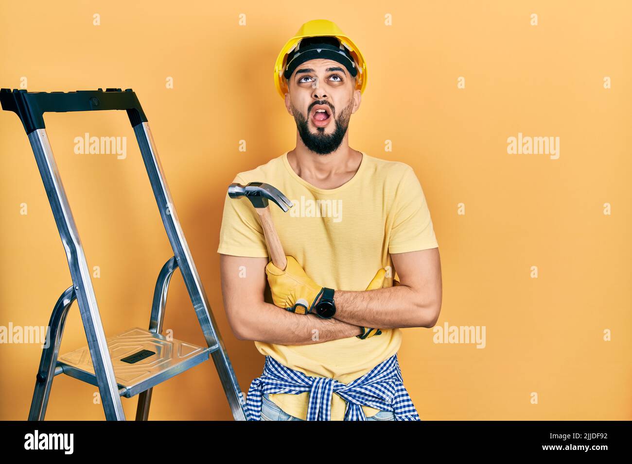 Handsome man with beard wearing hardhat holding hammer by stairs angry ...