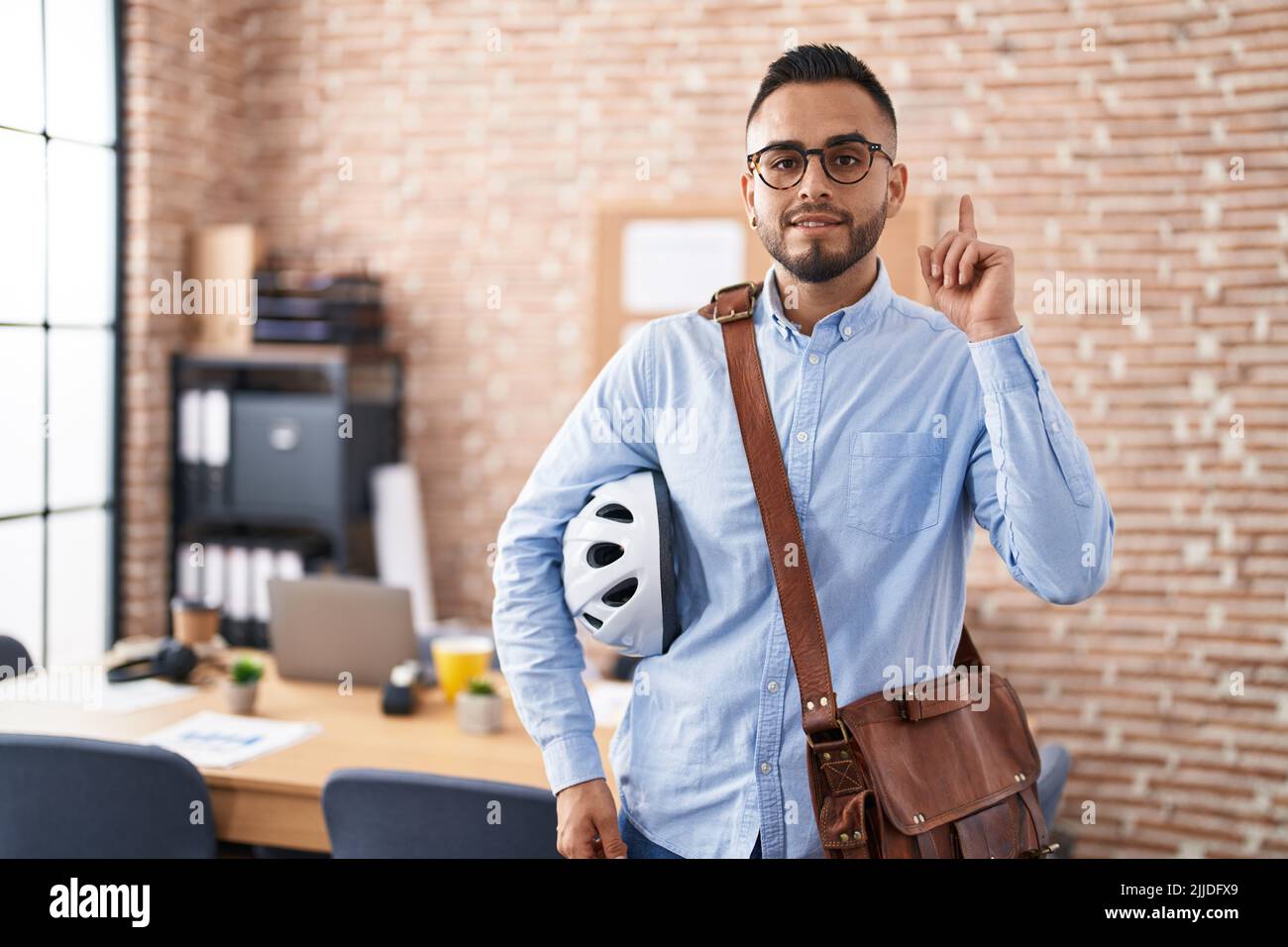 Young hispanic man working at the office holding bike helmet surprised ...