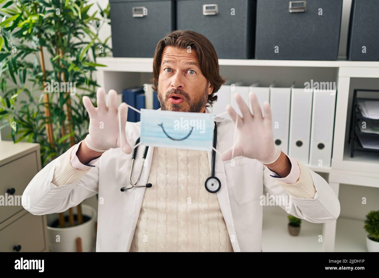 Handsome middle age doctor man wearing safety mask in shock face ...