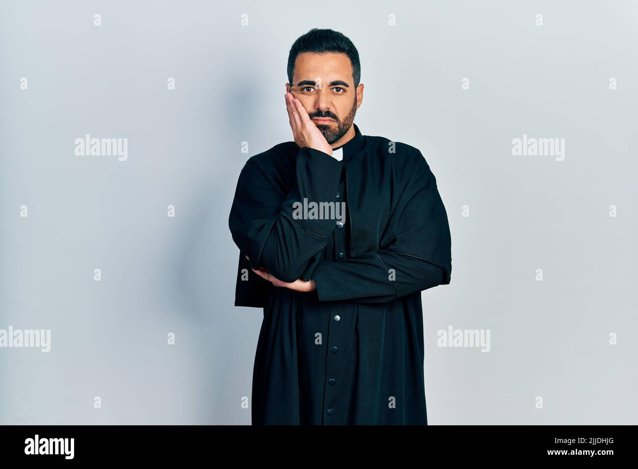 Handsome hispanic man with beard wearing catholic priest robe thinking ...