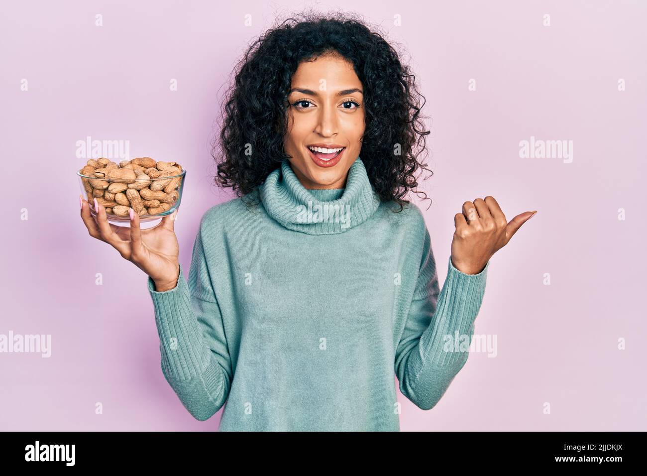 Young latin girl holding peanuts pointing thumb up to the side smiling ...