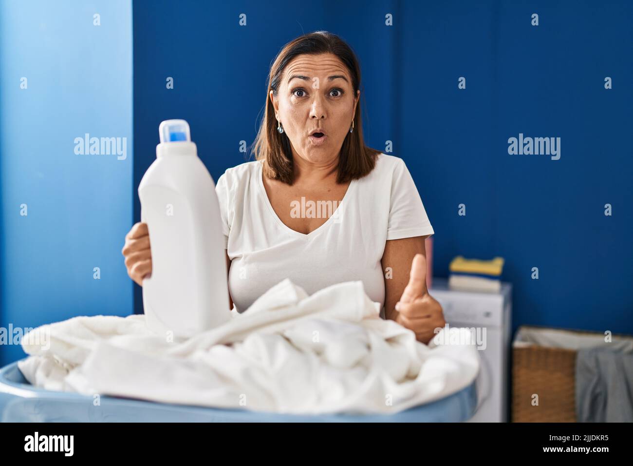 Middle age hispanic woman doing laundry holding detergent bottle scared and amazed with open ...