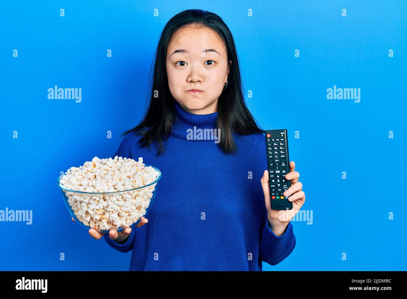 Young chinese girl eating popcorn using tv control puffing cheeks with ...