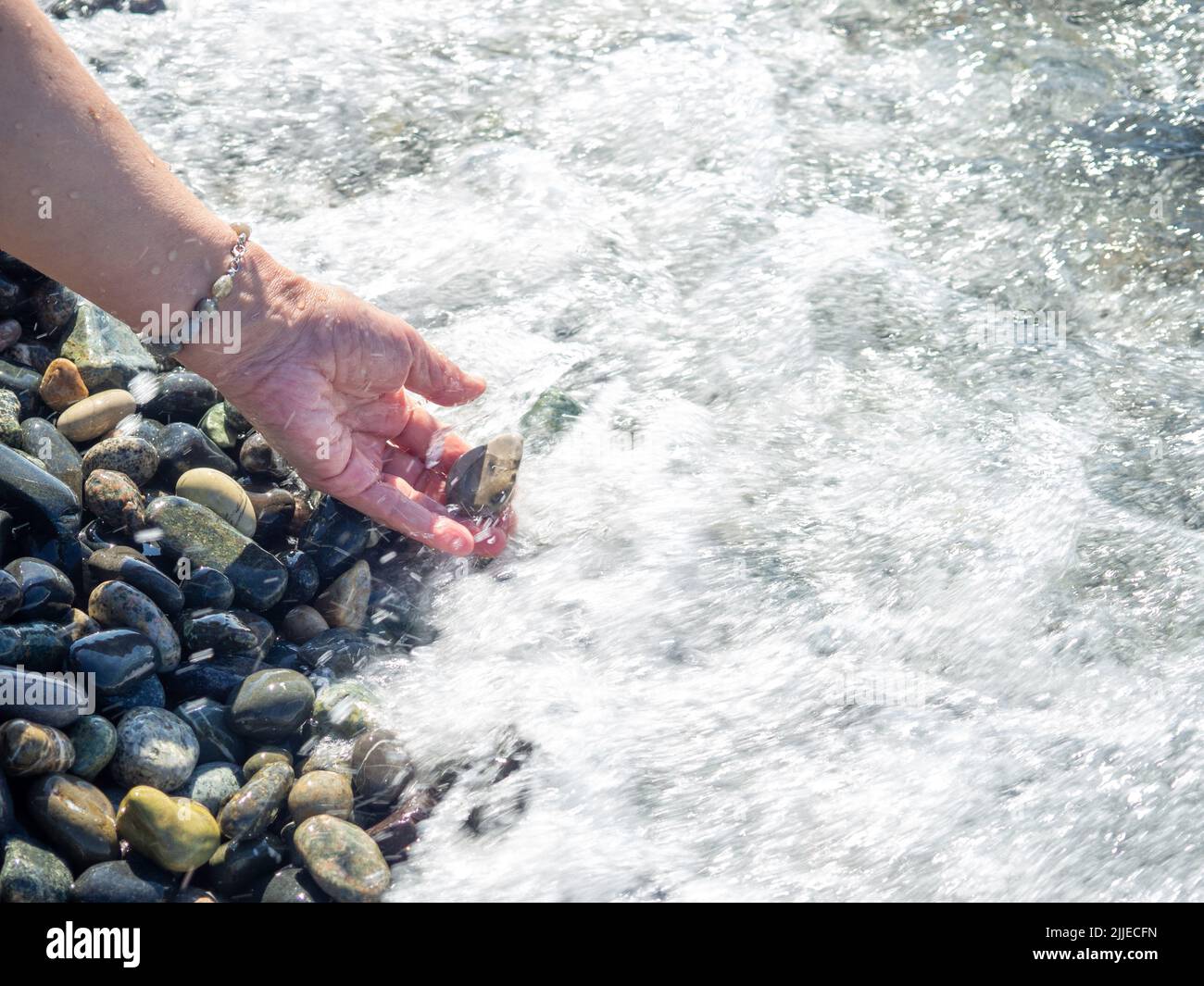 A hand with a bracelet made of stones holds a pebble near the water ...